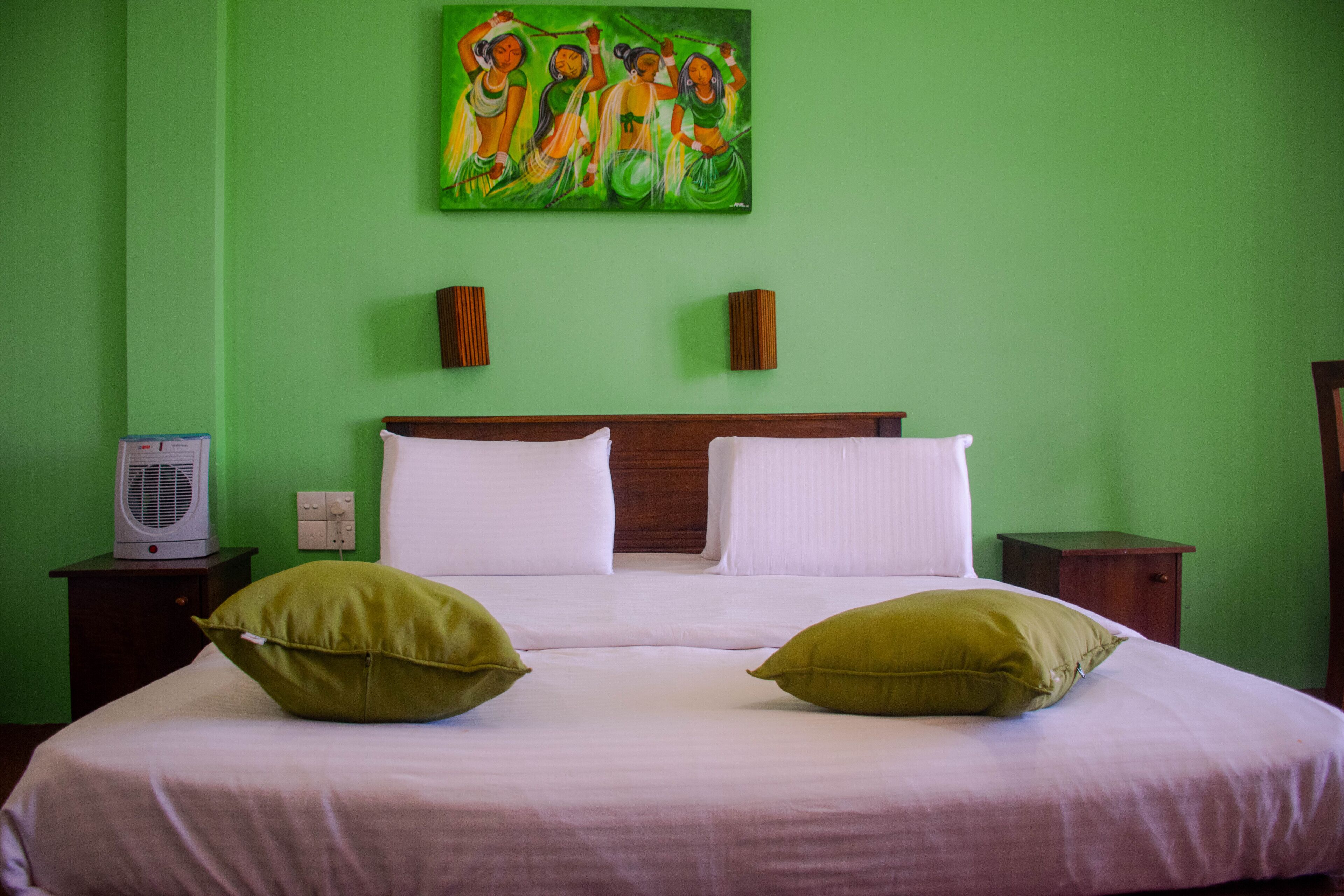 Quarto de hotel com uma cama grande, duas mesas de cabeceira, ar condicionado fixado na parede e uma parede verde com uma pintura.