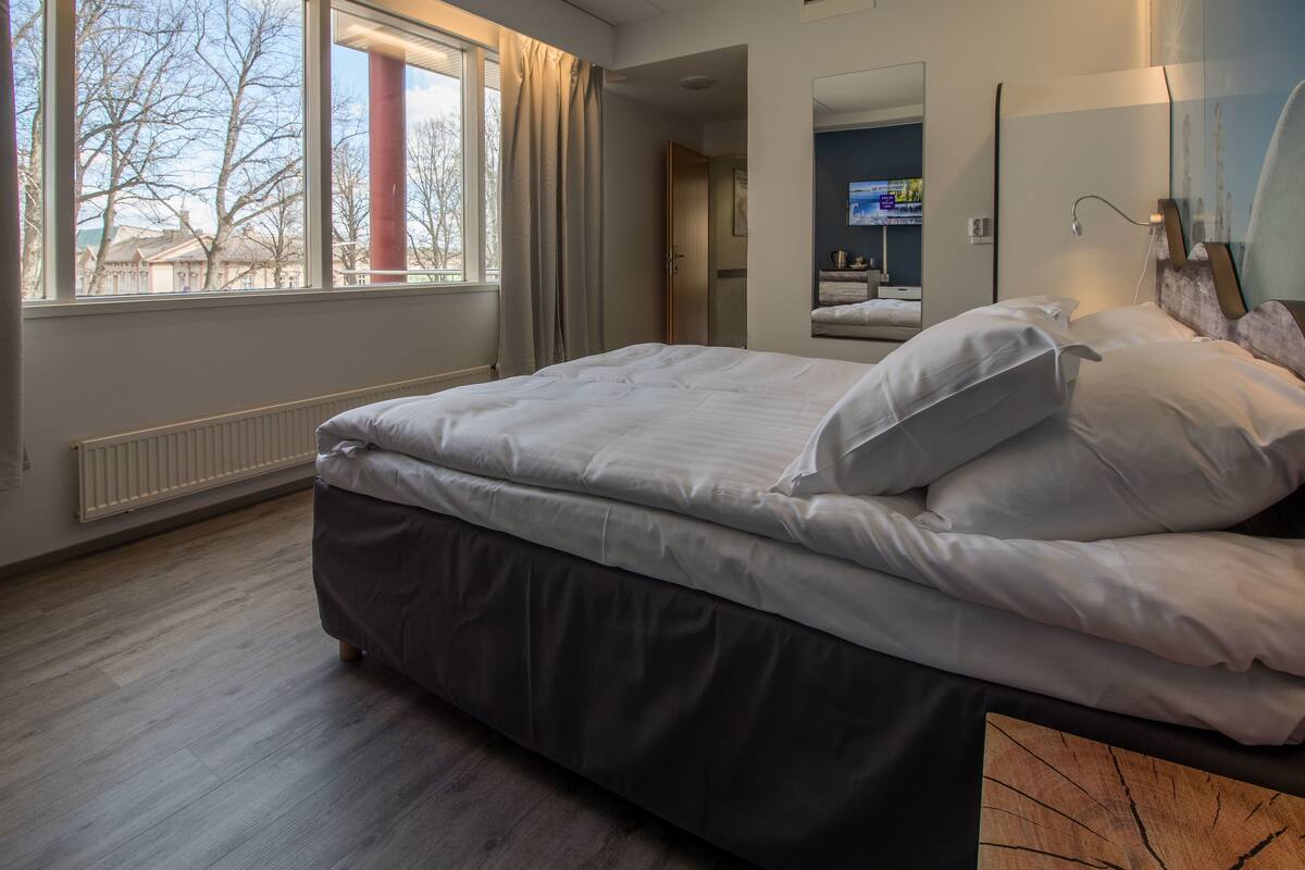 Quarto moderno com uma cama grande, janela com cortinas, radiador e espelho na parede.
