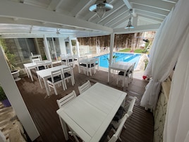 Uma área de refeições ao ar livre coberta, com mesas e cadeiras brancas, piscina ao fundo e uma parede de pedra.