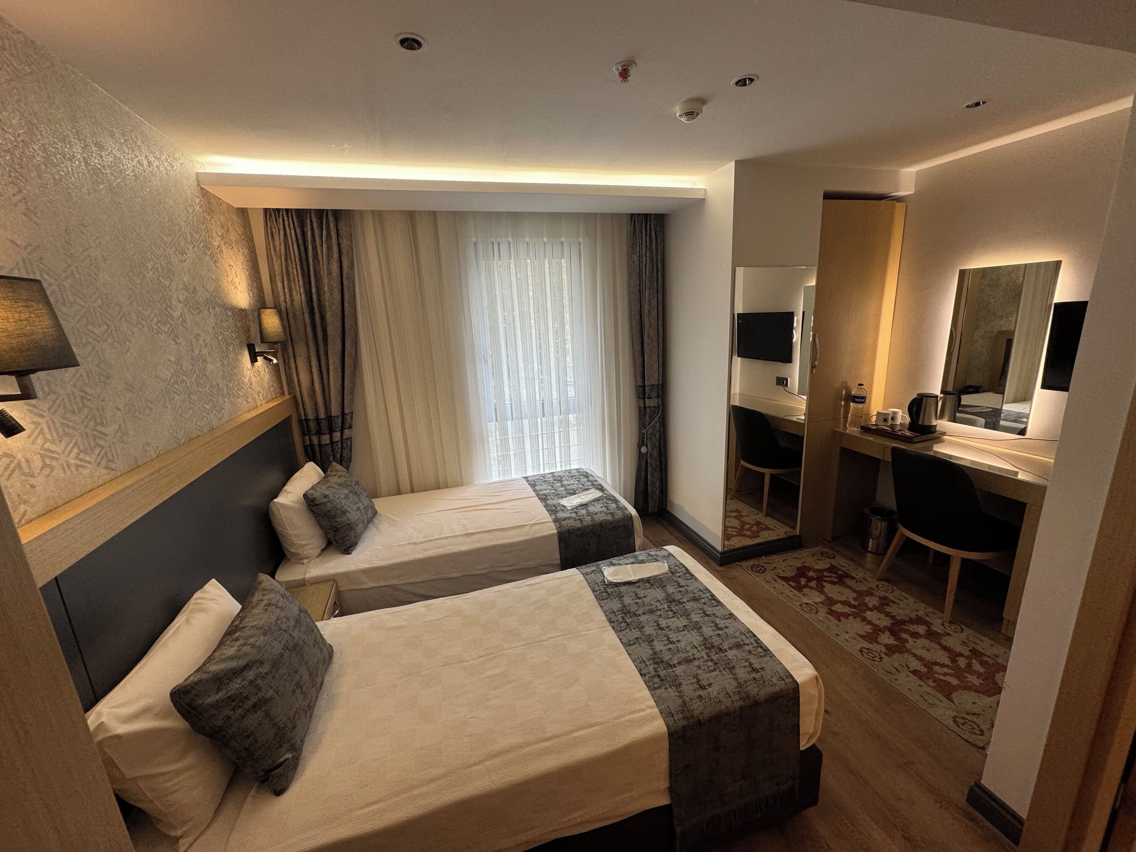 Quarto de hotel com duas camas, uma escrivaninha, uma cadeira, um espelho e uma televisão.