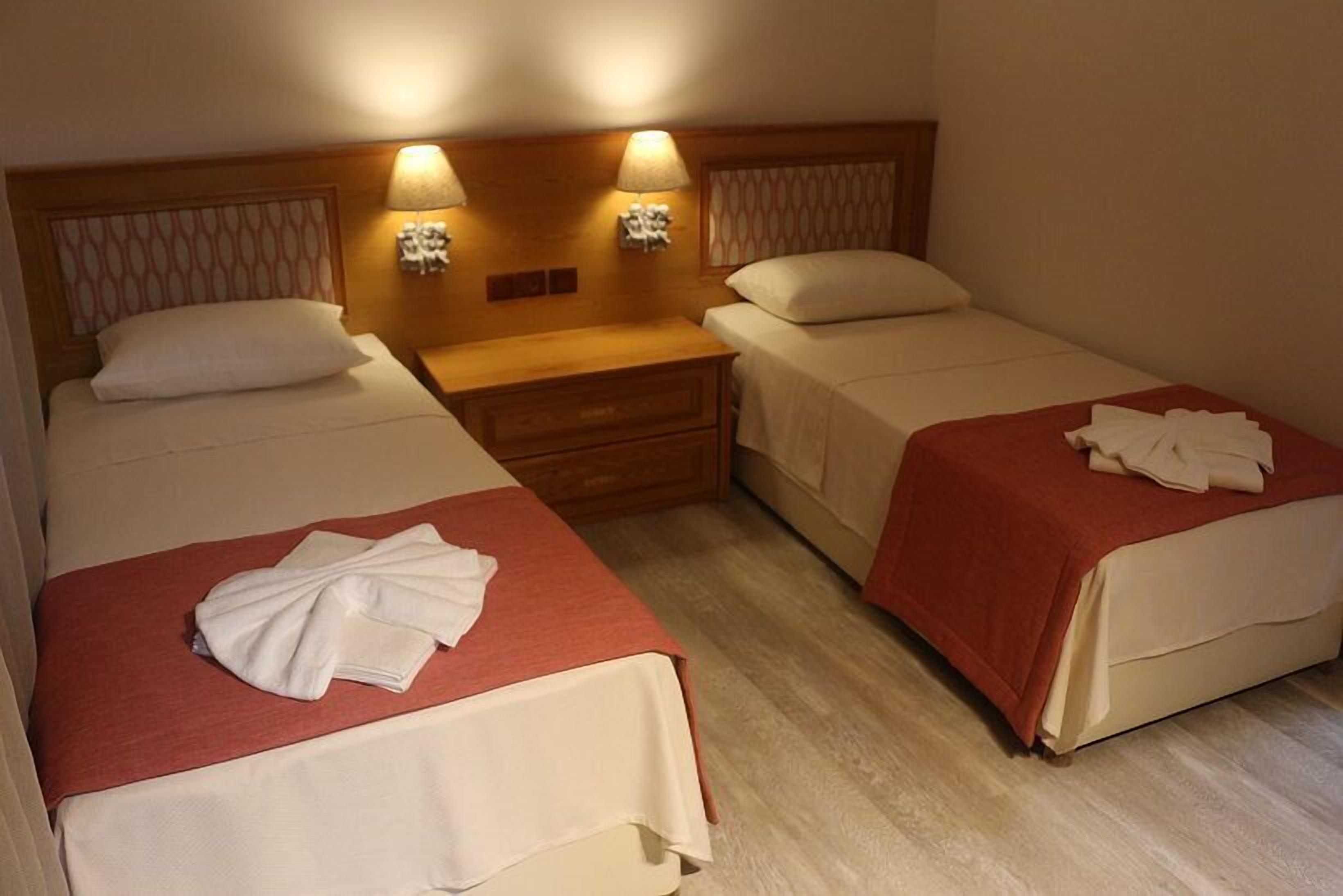 Quarto de hotel com duas camas de solteiro, uma mesinha de cabeceira de madeira e duas luminárias fixadas na parede.