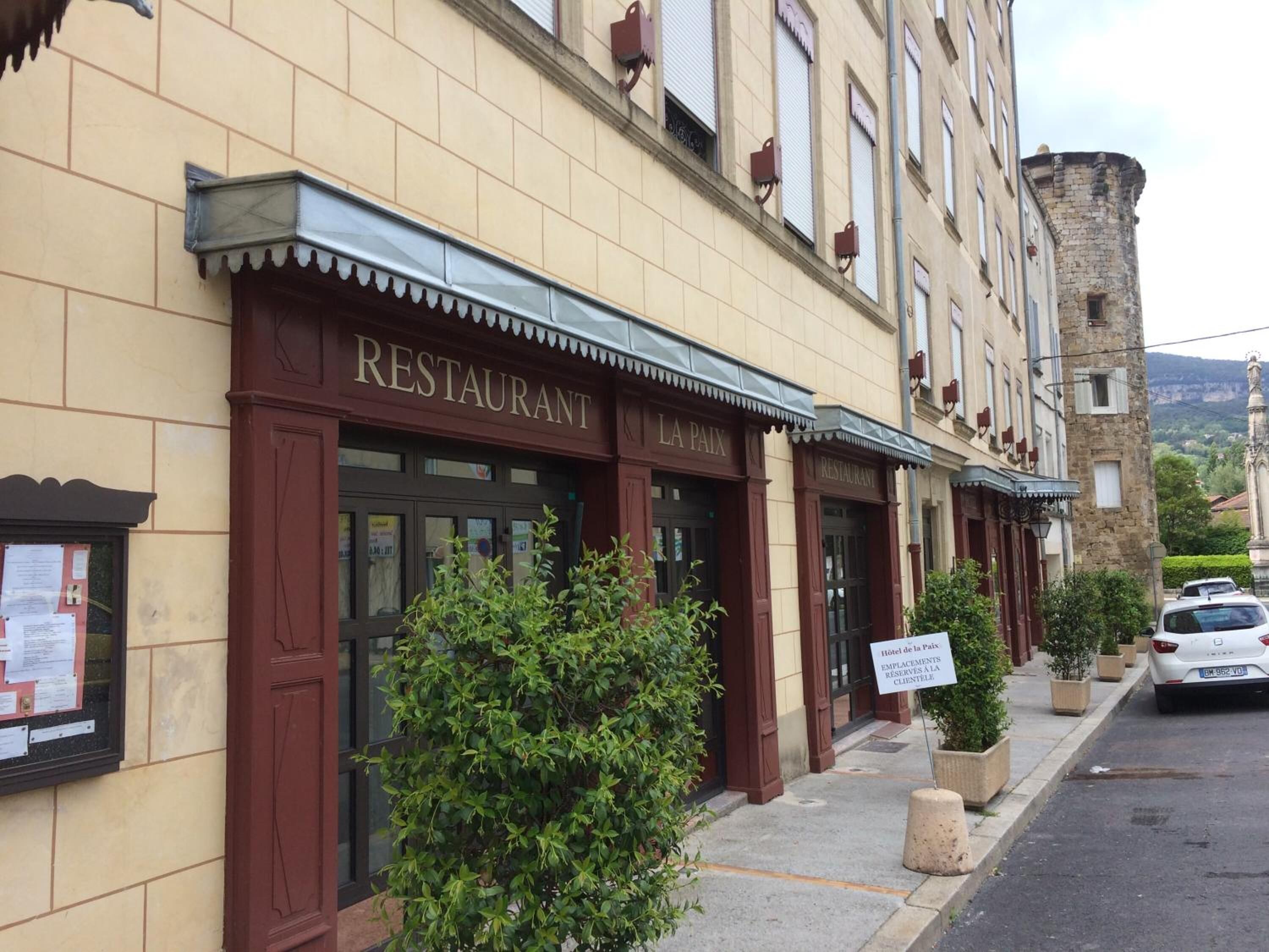 Um restaurante com fachada clássica, uma placa indicando ser o Hotel de la Paix, e uma torre de pedra ao fundo.