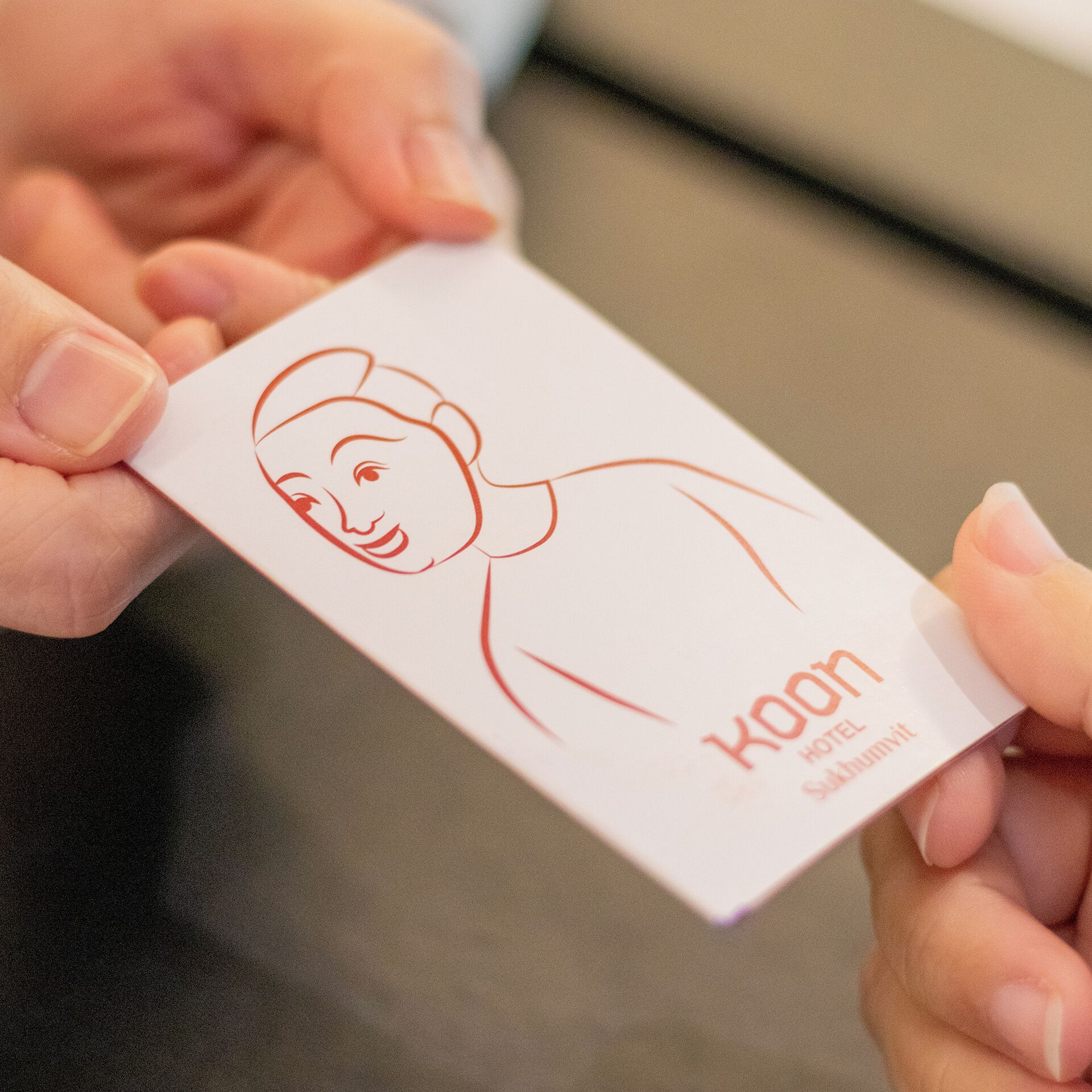 Uma mão segurando um cartão de visita com um desenho simples de uma pessoa e o texto 'Koon Hotel' e 'Sukhumvit'.