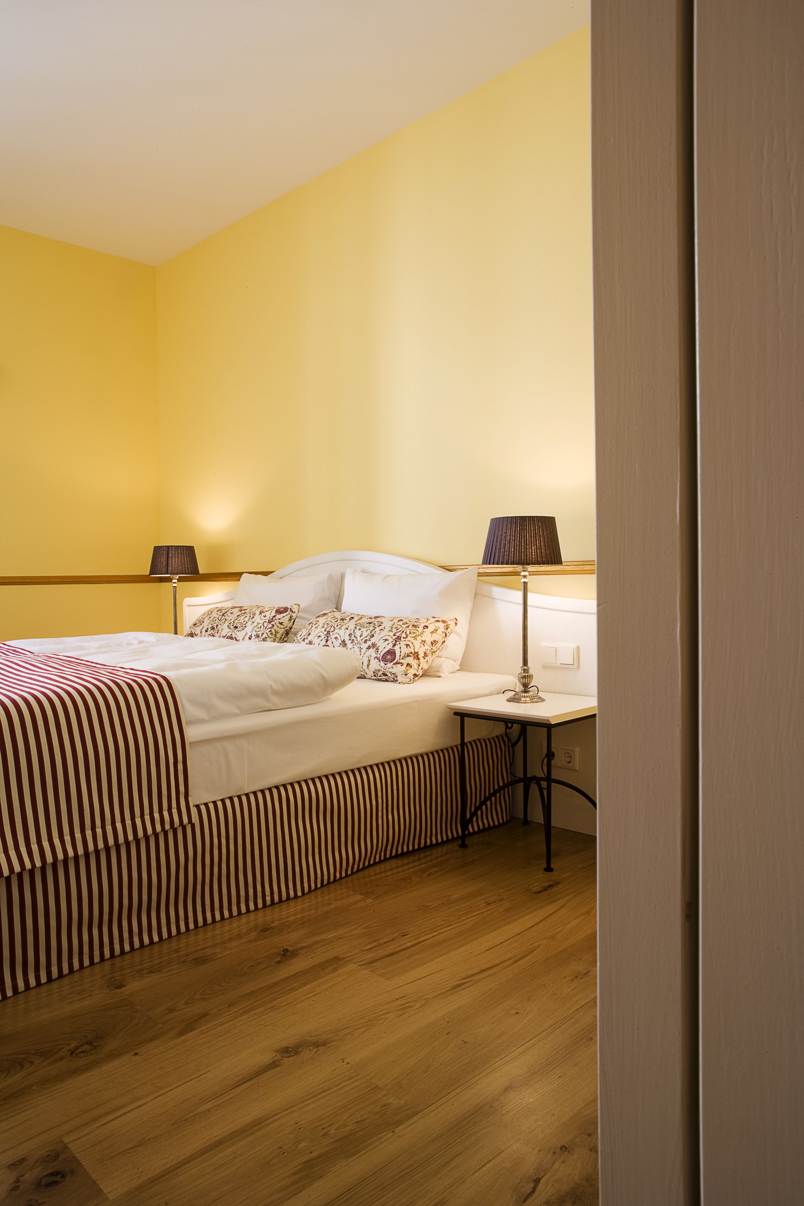 Quarto de hotel com cama, luminárias laterais e piso de madeira.