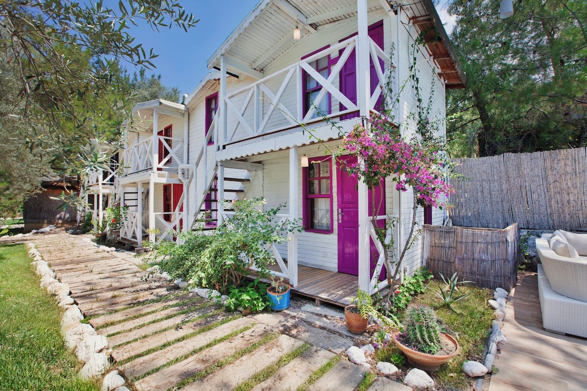 Uma casa branca com detalhes em roxo, um deck de madeira e um jardim com diversas plantas.
