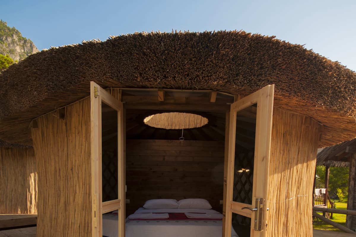 Uma cabana de madeira com telhado de palha, uma cama com lençóis brancos e um deck de madeira.