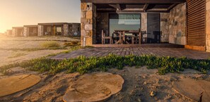 Uma cabana rústica com deck de madeira, paredes de pedra e uma grande janela com vista para uma área gramada.