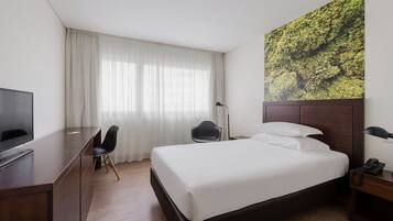 Quarto de hotel com cama, escrivaninha, cadeira, televisão, luminária e um quadro na parede.