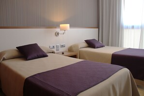 Quarto de hotel com duas camas, cada uma com almofada roxa e edredom bege. Há uma luminária na parede à esquerda da imagem, e uma janela com cortinas à direita.