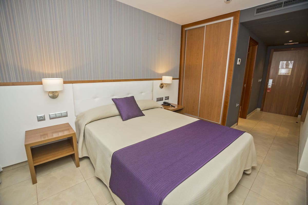Quarto de hotel com cama, mesa de cabeceira, luminária na parede e guarda-roupa.