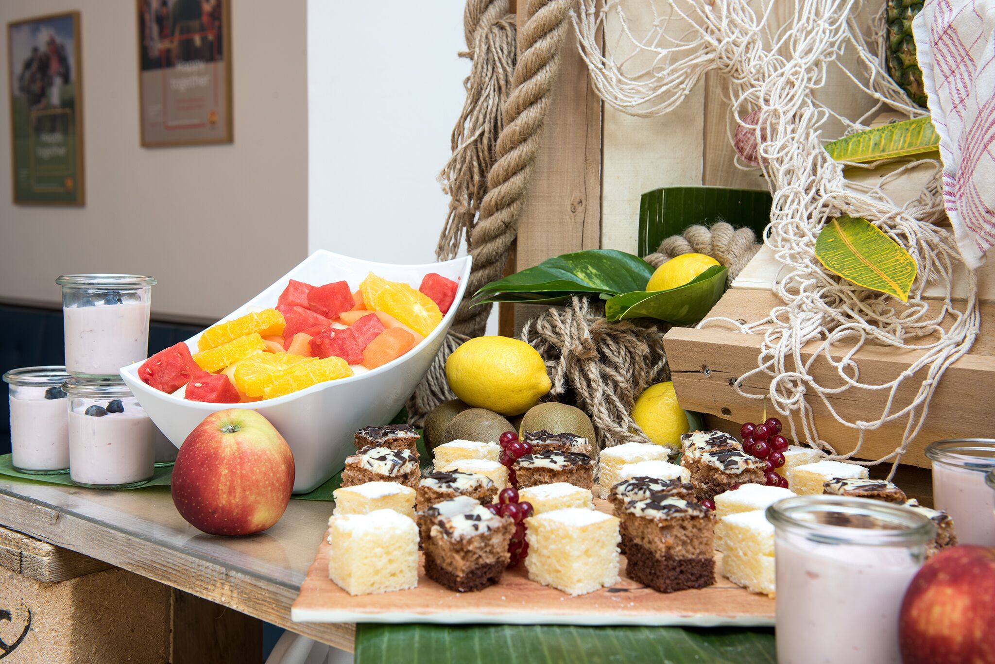Uma mesa com diversos alimentos, incluindo uma tigela de frutas, um prato de sobremesas e potes com o que parece ser iogurte ou produto lácteo similar.