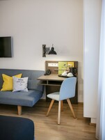 Quarto de hotel moderno com sofá, escrivaninha com telefone e chaleira, uma cadeira e uma TV de tela plana fixada na parede.