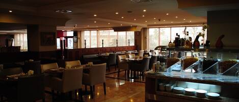 Restaurante com várias mesas e cadeiras, um balcão de buffet com pratos e tigelas, e grandes janelas que permitem a entrada de luz natural.