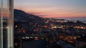 Vista urbana ao entardecer, com a costa, edifícios e uma colina ao longe.