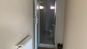 Um corredor estreito com um radiador, uma porta de acesso a um banheiro e um par de sapatos perto da porta.