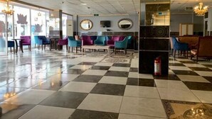 Um lobby com piso xadrez, várias áreas de estar com cadeiras, um extintor de incêndio e um lustre.