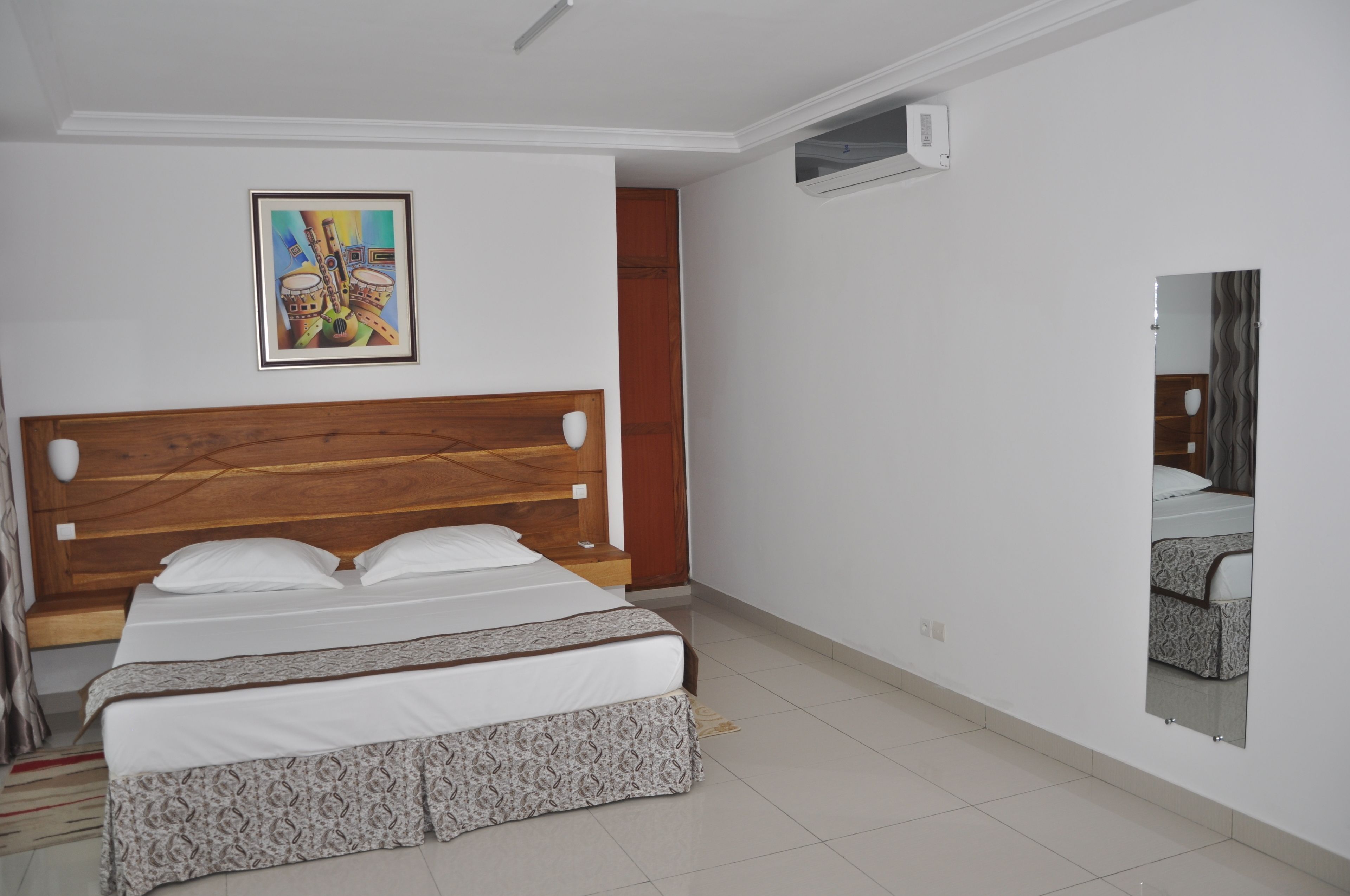 Quarto com cabeceira de madeira, cama com roupa de cama branca, saia de cama estampada, pintura na parede e um espelho refletindo a cama.