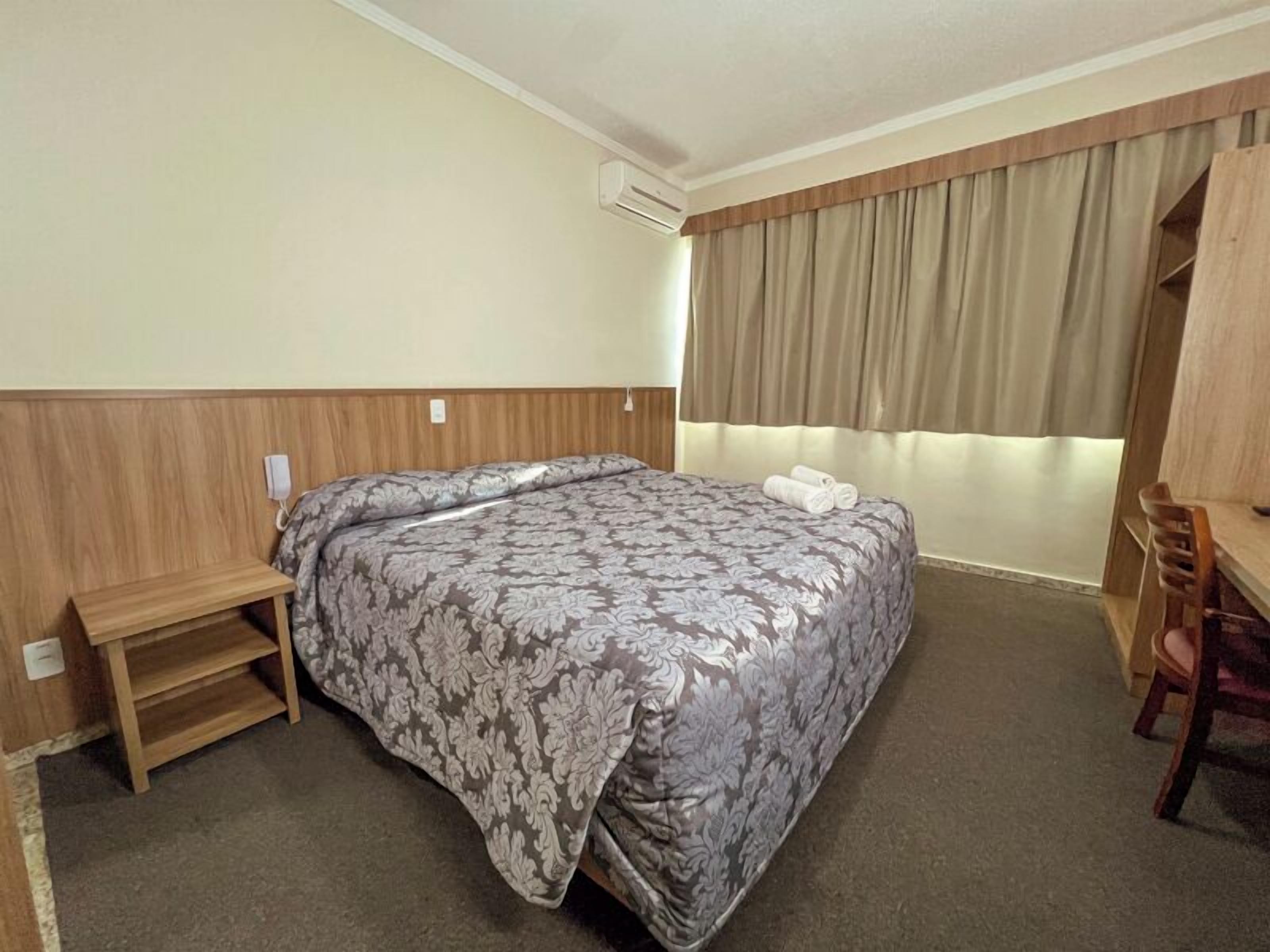 Quarto de hotel com uma cama, uma mesinha de cabeceira de madeira, uma escrivaninha e uma cadeira.
