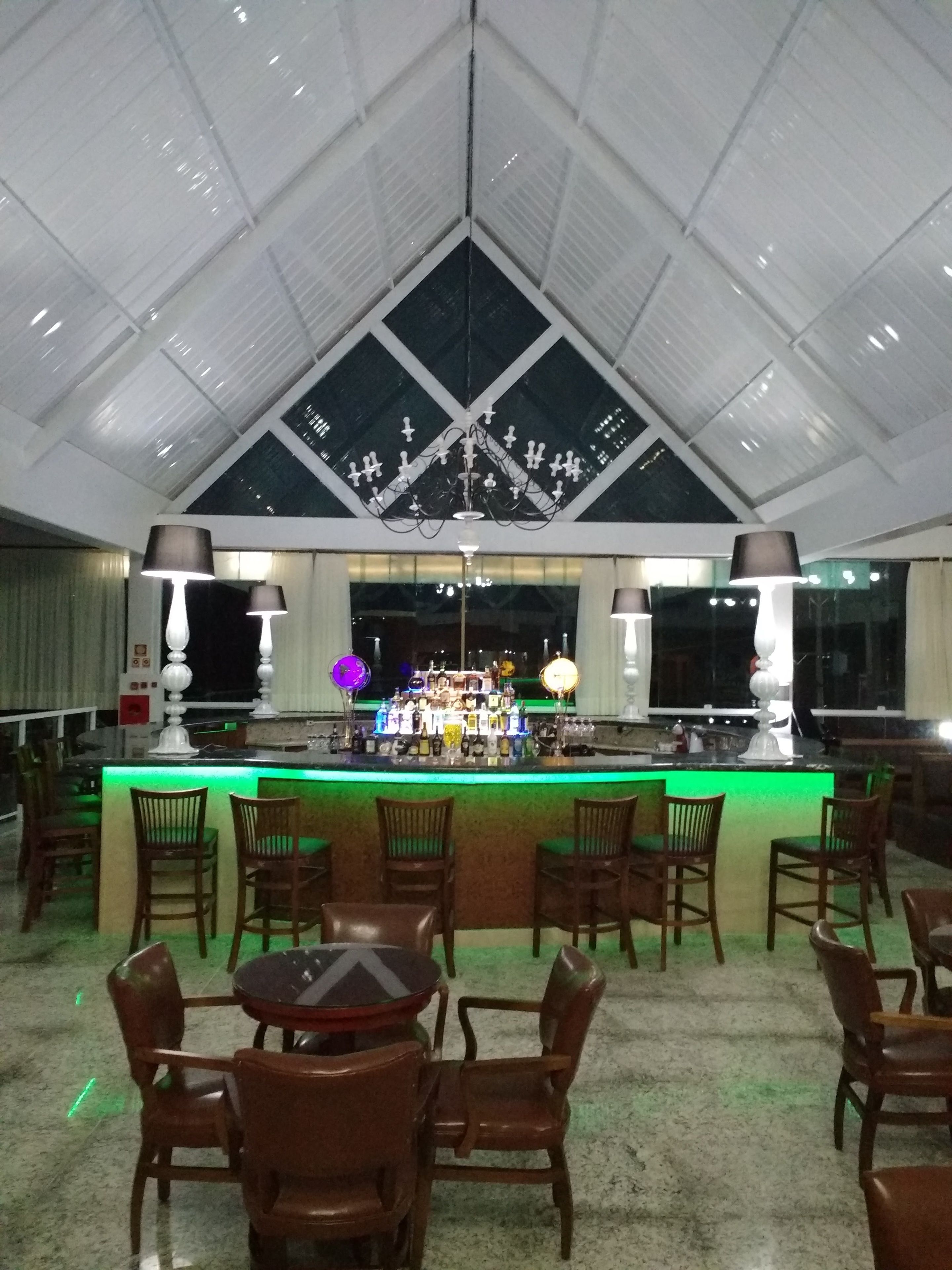Uma área de bar com teto alto e um candelabro. Há várias cadeiras e mesas ao redor do balcão.