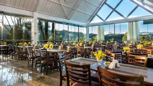 Um restaurante espaçoso com tetos altos e grandes janelas que oferecem vista para o exterior. As mesas estão bem organizadas com cadeiras, e cada uma tem um vaso com flores amarelas.