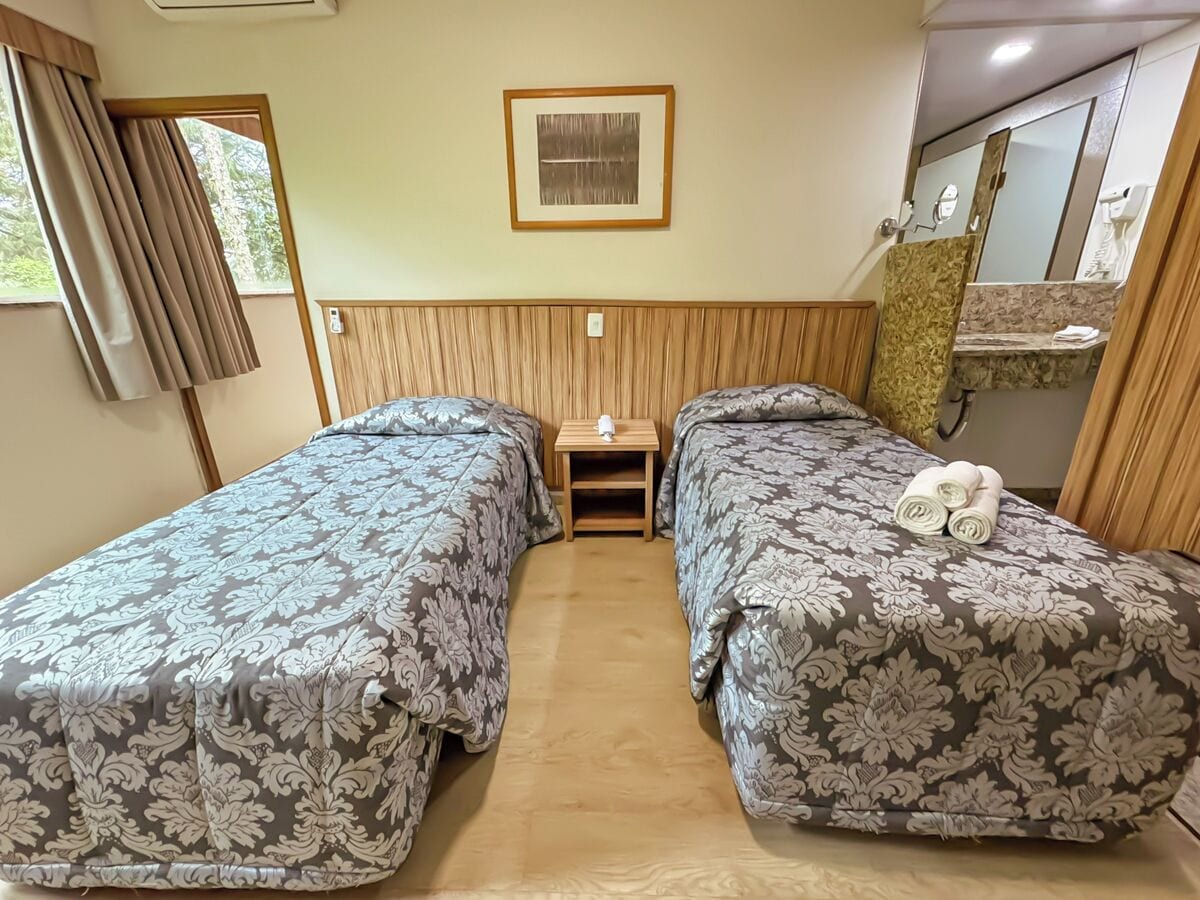 Quarto de hotel com duas camas de solteiro, cabeceira de madeira, mesinha de cabeceira com abajur e espelho de banheiro.