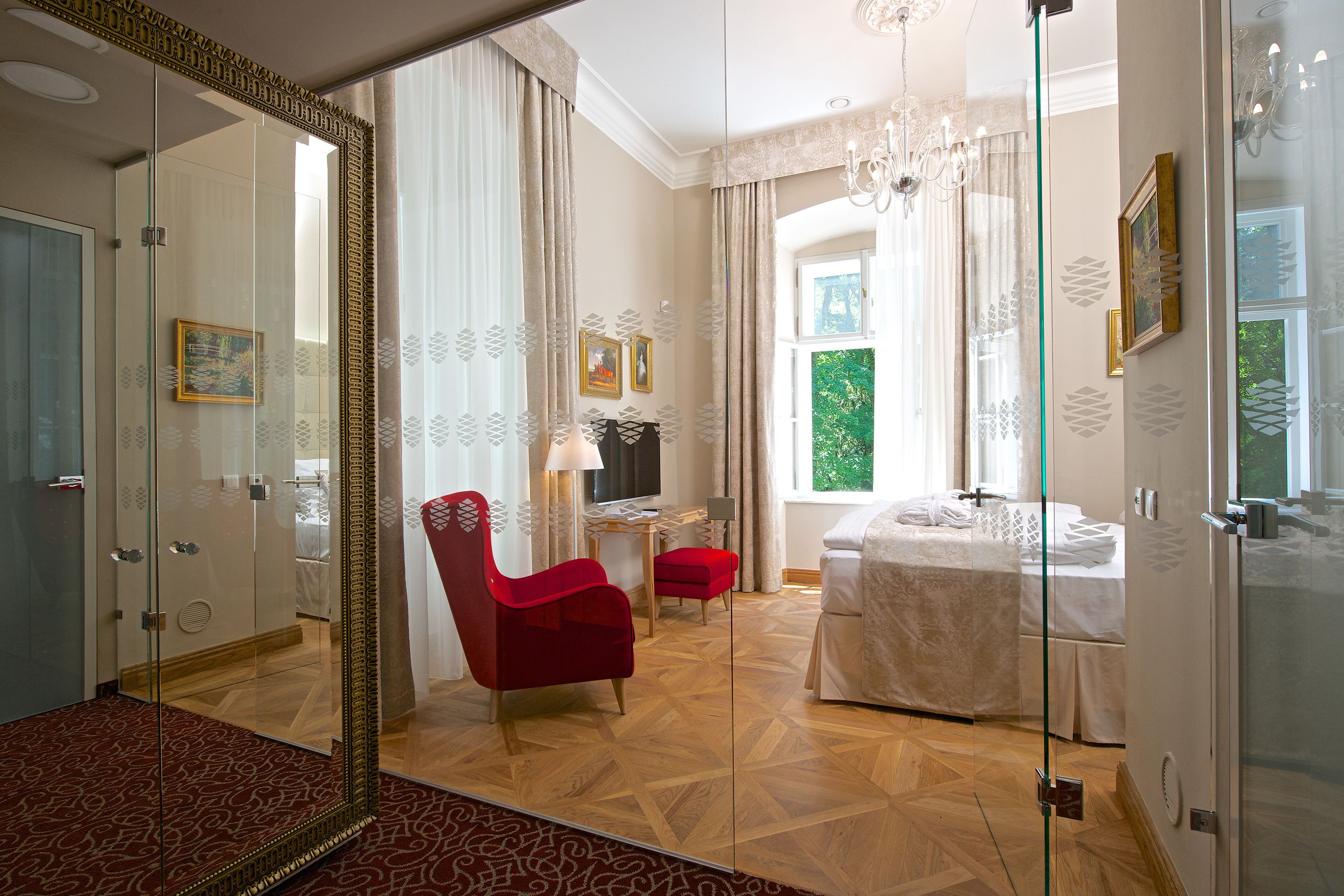 Quarto de hotel com um espelho grande, uma cadeira vermelha, uma cama e uma escrivaninha com abajur.