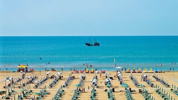 Uma praia com fileiras de espreguiçadeiras e guarda-sóis, um barco na água e um céu azul límpido.