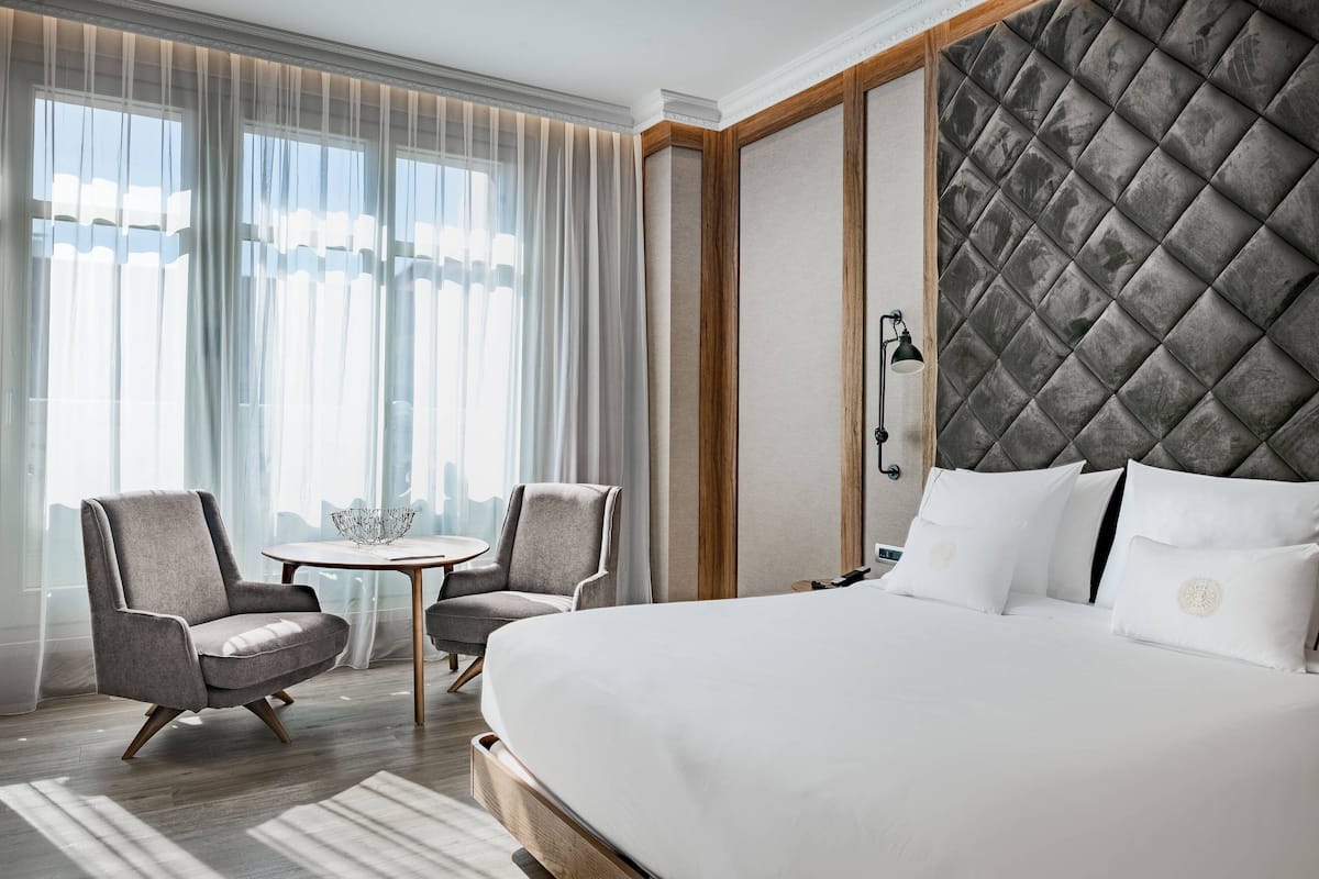 Quarto de hotel com uma cama grande, duas poltronas, uma mesinha redonda e uma cabeceira decorativa.