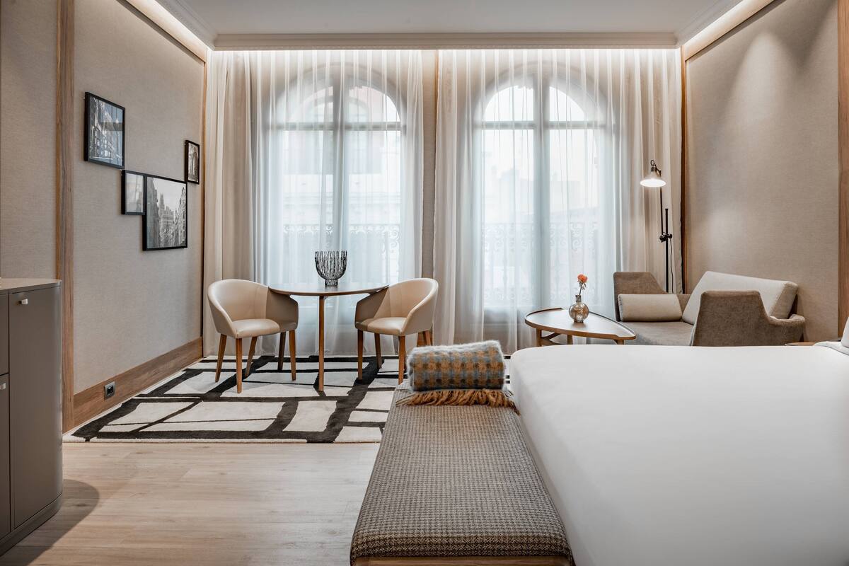 Quarto de hotel com uma cama, duas cadeiras, uma mesinha e um abajur.