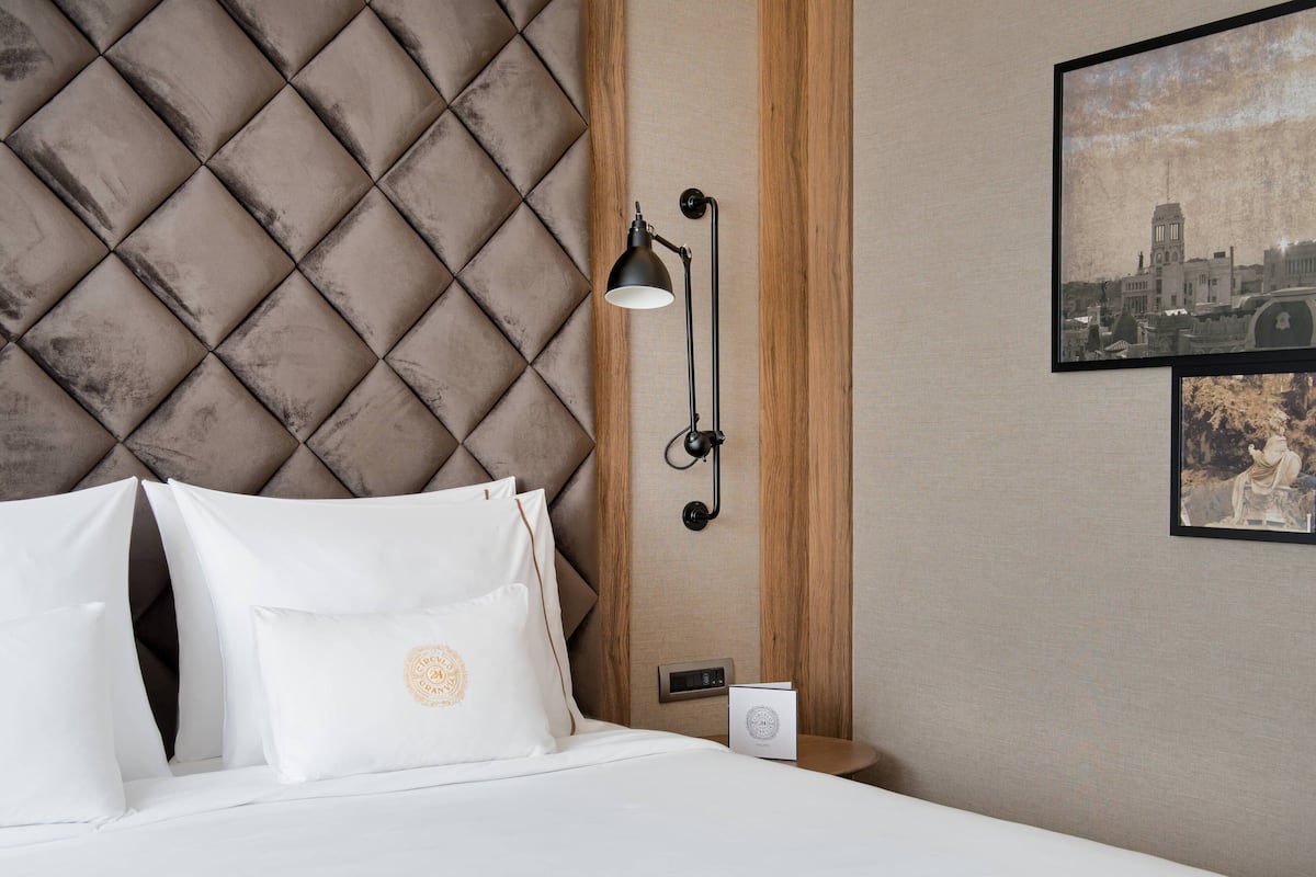 Quarto de hotel com cabeceira estofada, mesa de cabeceira com abajur e quadro na parede.