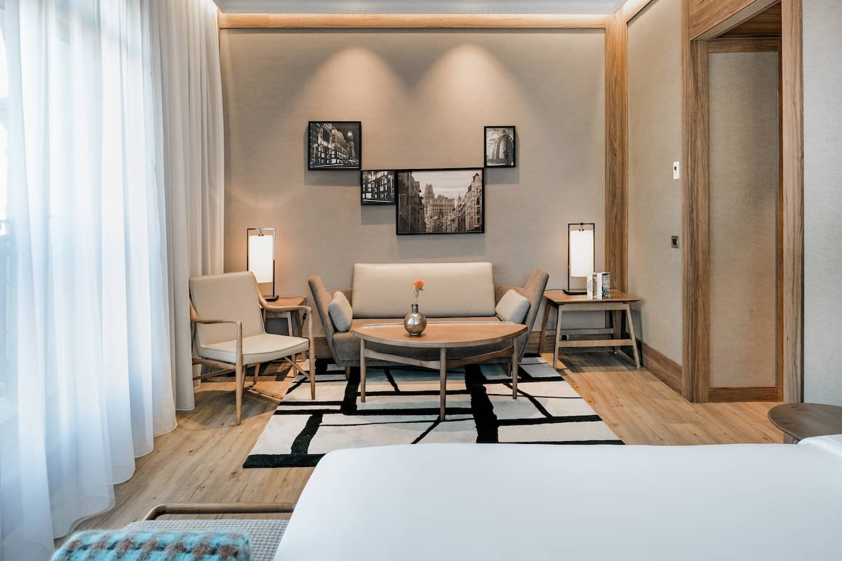 Quarto de hotel moderno com uma cama, um sofá, duas poltronas, uma mesa de centro e uma escrivaninha com uma luminária. Há quadros nas paredes e uma grande janela com cortinas.