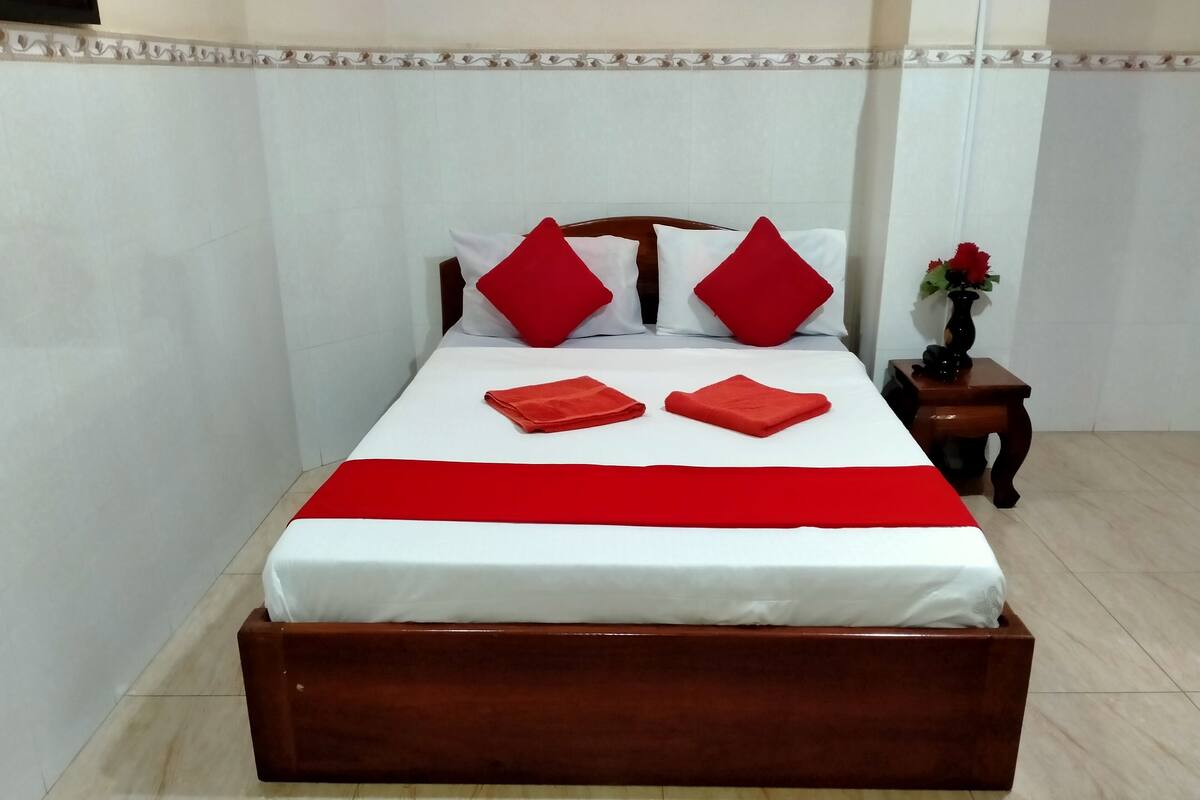 Cama bem arrumada com colcha vermelha e branca, duas toalhas vermelhas e cabeceira de madeira.