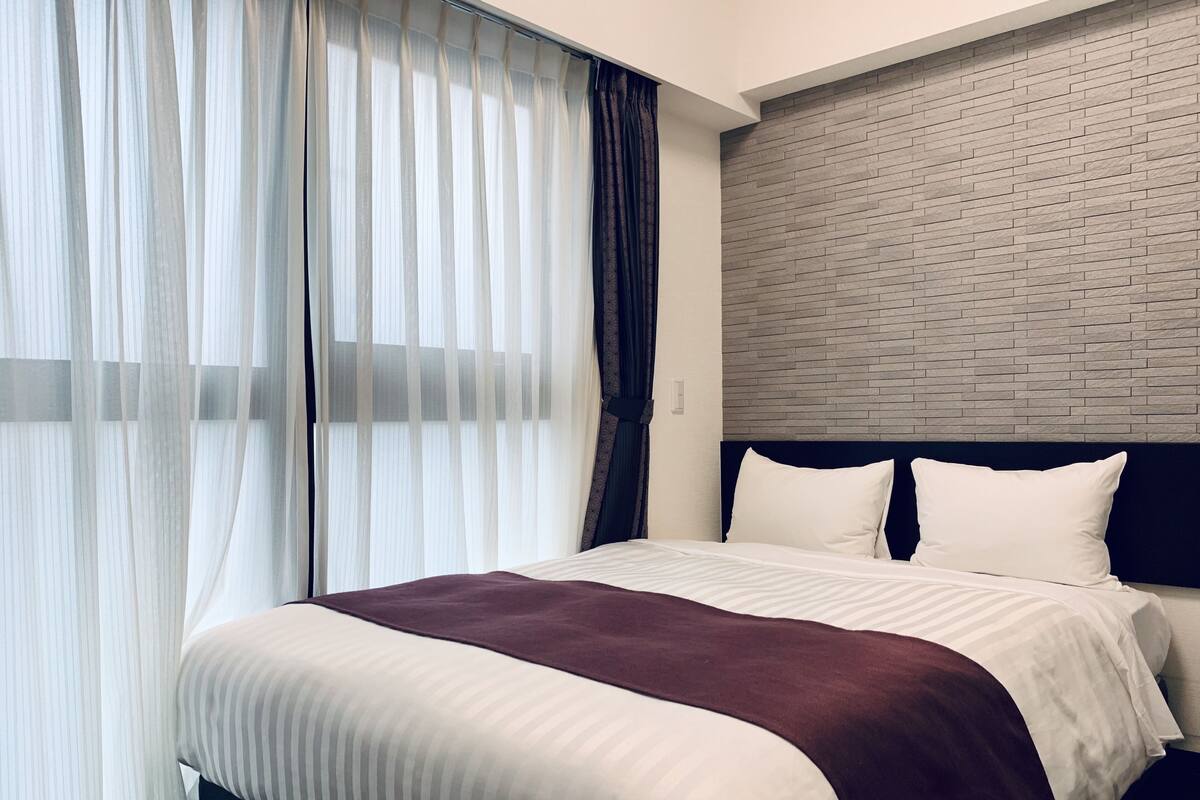 Quarto de hotel moderno com uma cama grande, cabeceira em padrão de tijolos e cortinas leves.