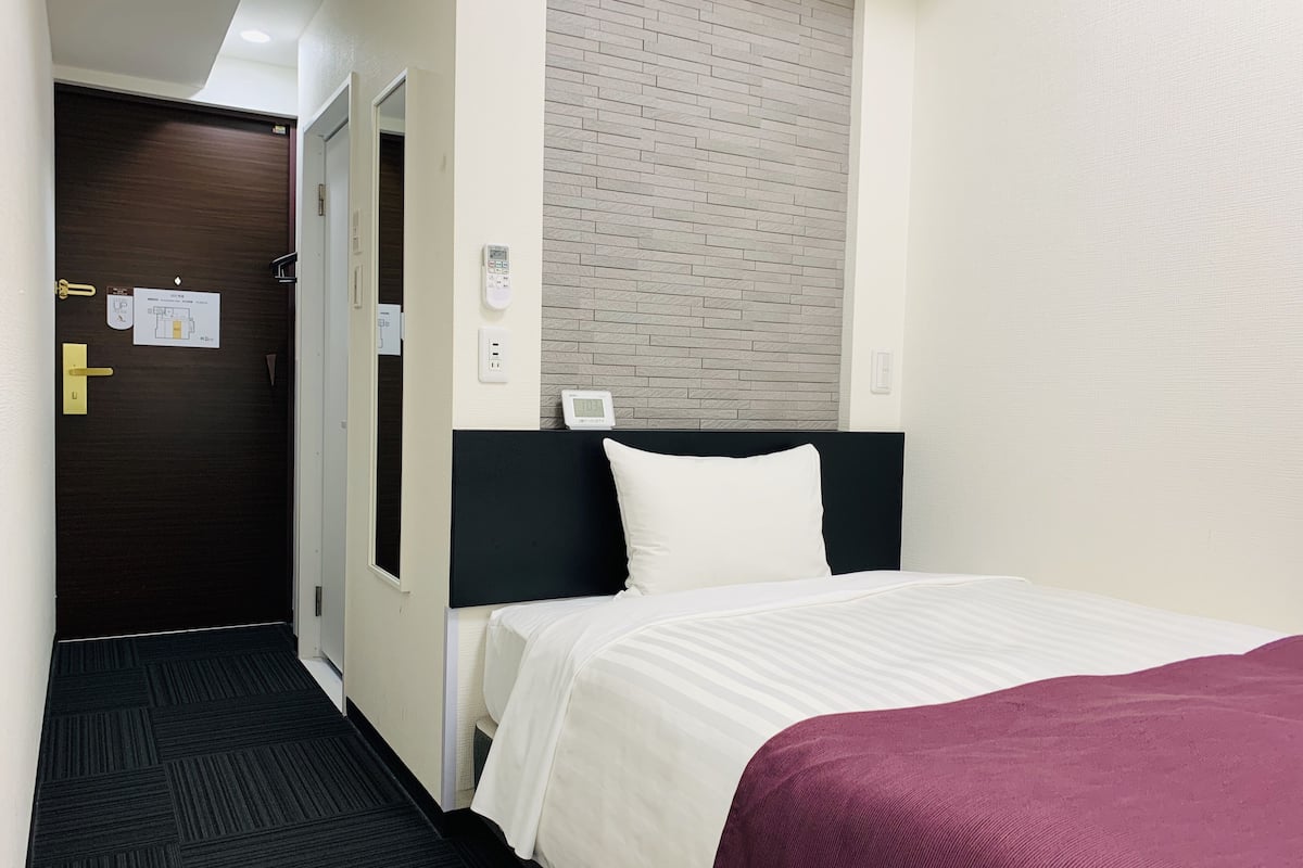 Quarto de hotel com cama, porta de madeira escura, parede branca e parede de destaque com padrão de tijolos, além de um relógio digital.