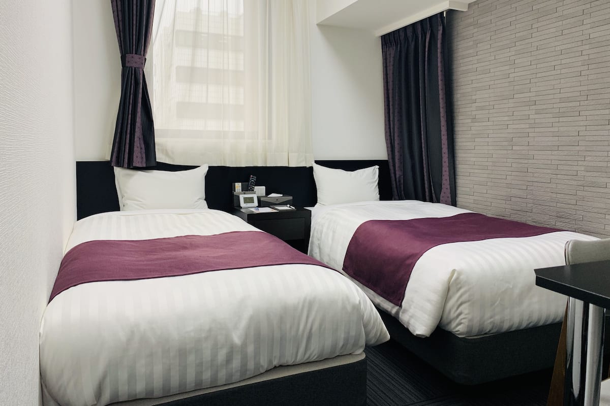 Quarto de hotel com duas camas, uma mesinha de cabeceira, uma escrivaninha e parede com padrão de tijolos.