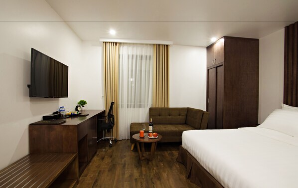 Quarto de hotel com uma cama, uma escrivaninha, uma cadeira, uma mesinha com uma garrafa e fruta, uma televisão e um guarda-roupa.