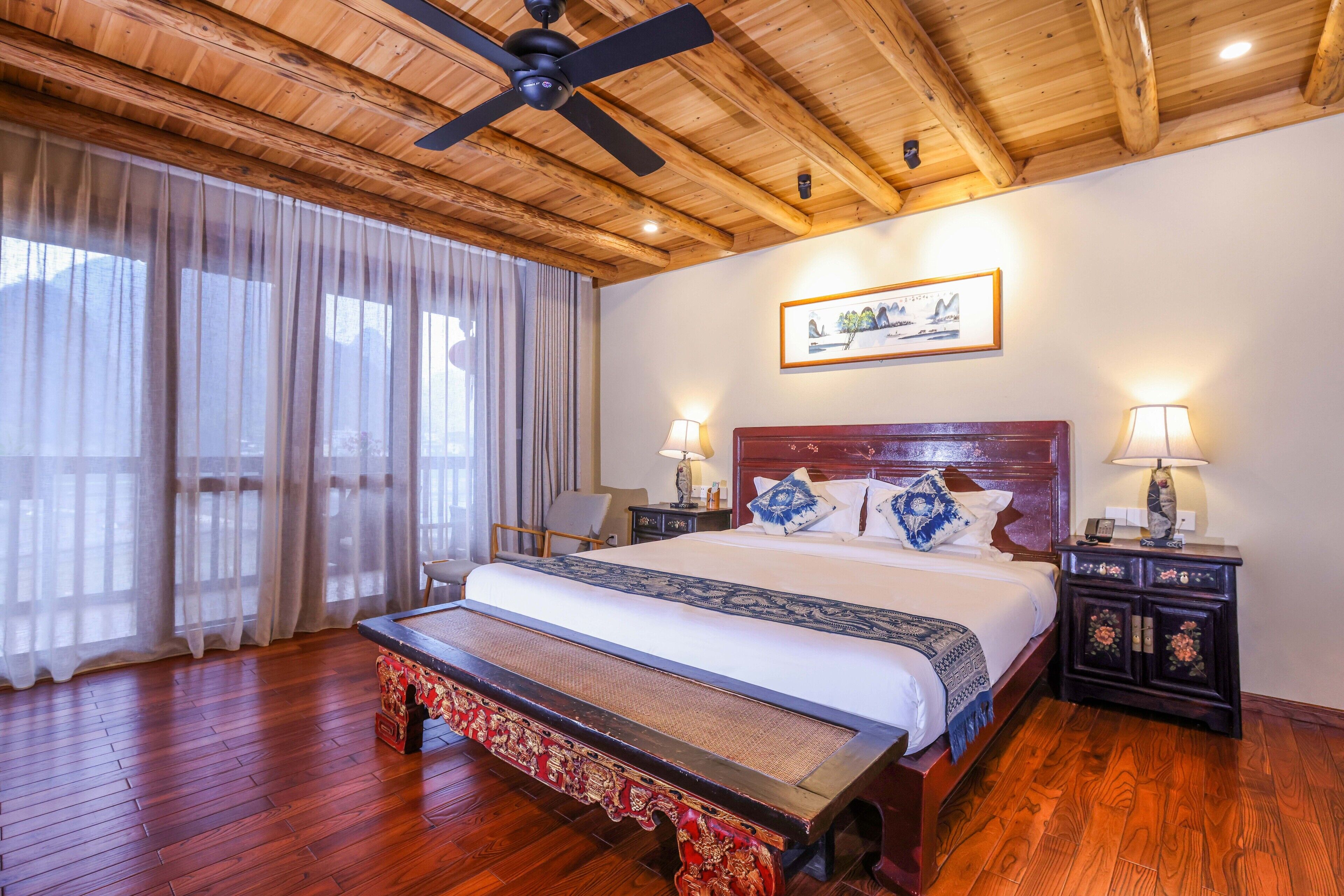 Um quarto espaçoso com uma cama grande, piso de madeira, ventoinha no teto e vista para o exterior por grandes janelas.