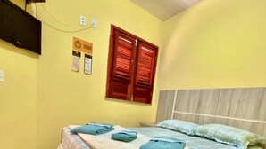Quarto com cama, cabeceira de madeira, janela com persianas e televisão fixada na parede.