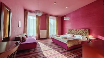 Quarto de hotel com parede rosa, uma cama com cabeceira floral, sofá rosa, uma mesinha e uma cadeira.
