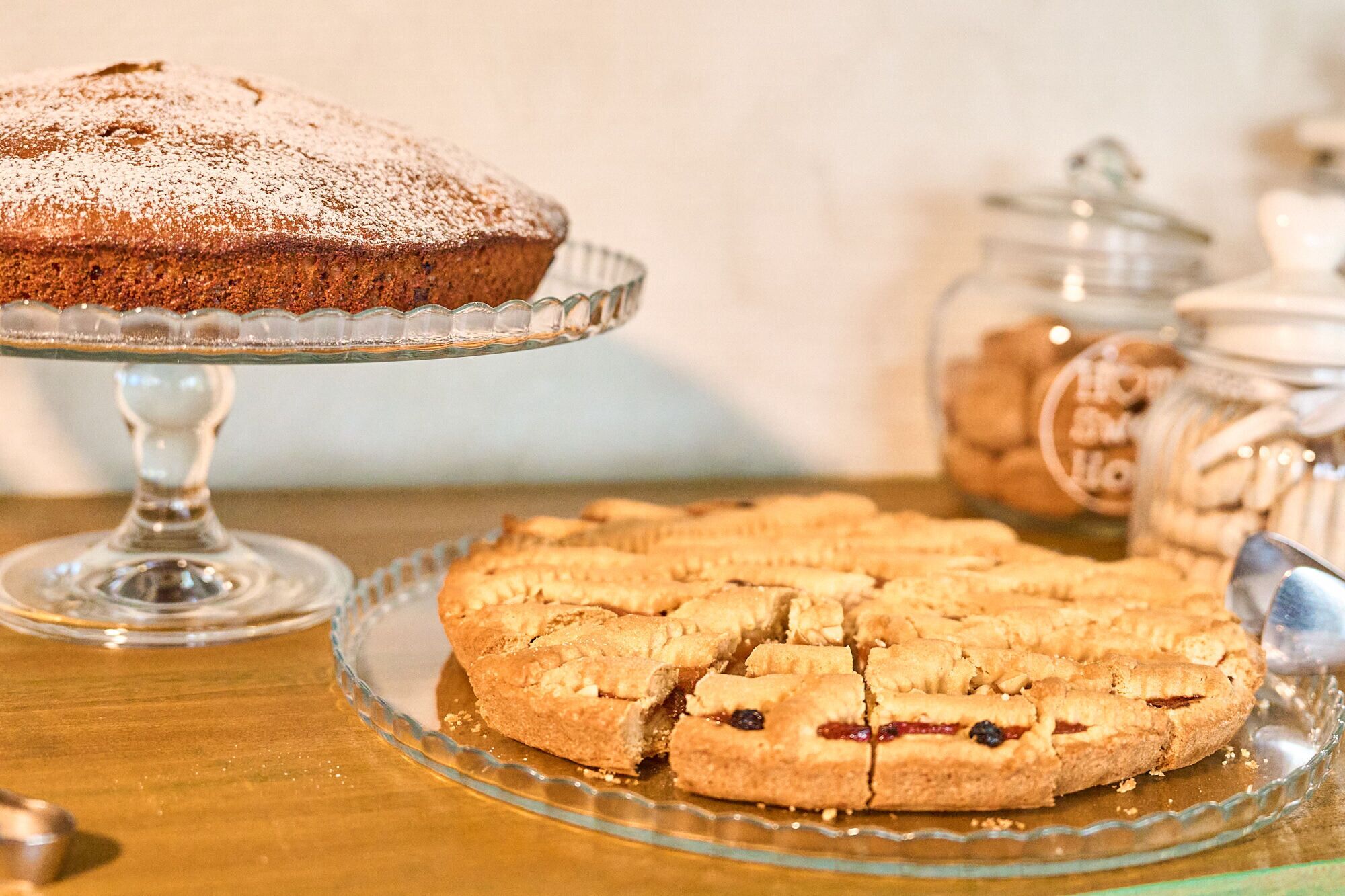 Um bolo sobre um suporte de vidro e uma bandeja com biscoitos cortados em uma mesa de madeira.
