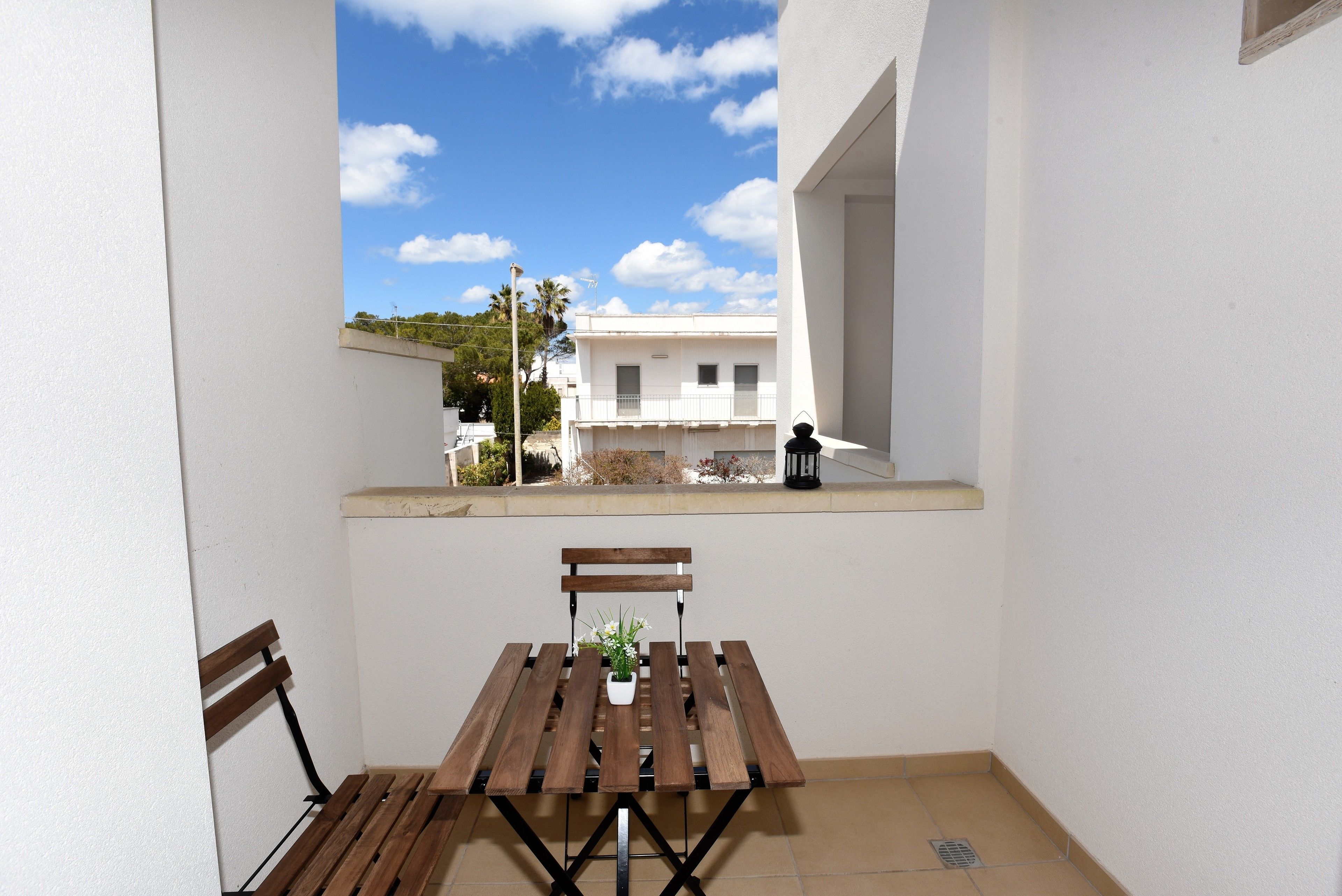 Uma varanda com mesa e banco de madeira, um vaso com planta e vista para um edifício branco sob céu azul.