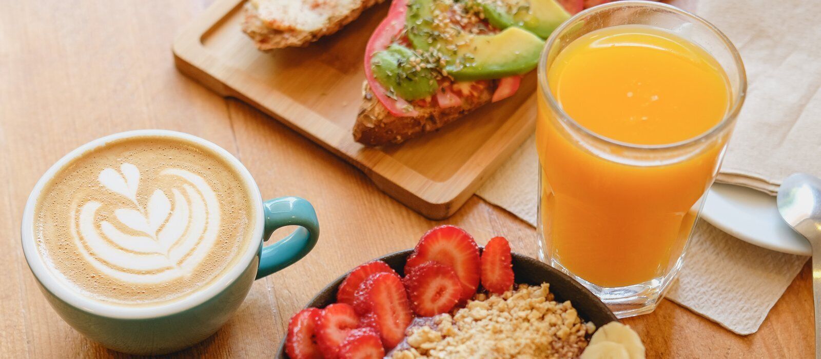 Uma mesa de café da manhã com xícara de café, torrada com abacate e tomate, uma tigela de morangos e granola, e um copo de suco de laranja.