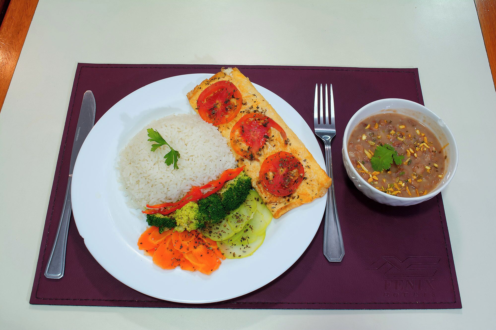 Um prato com arroz, legumes e uma fatia de pizza sobre uma mesa, com um garfo e uma tigela de sopa.