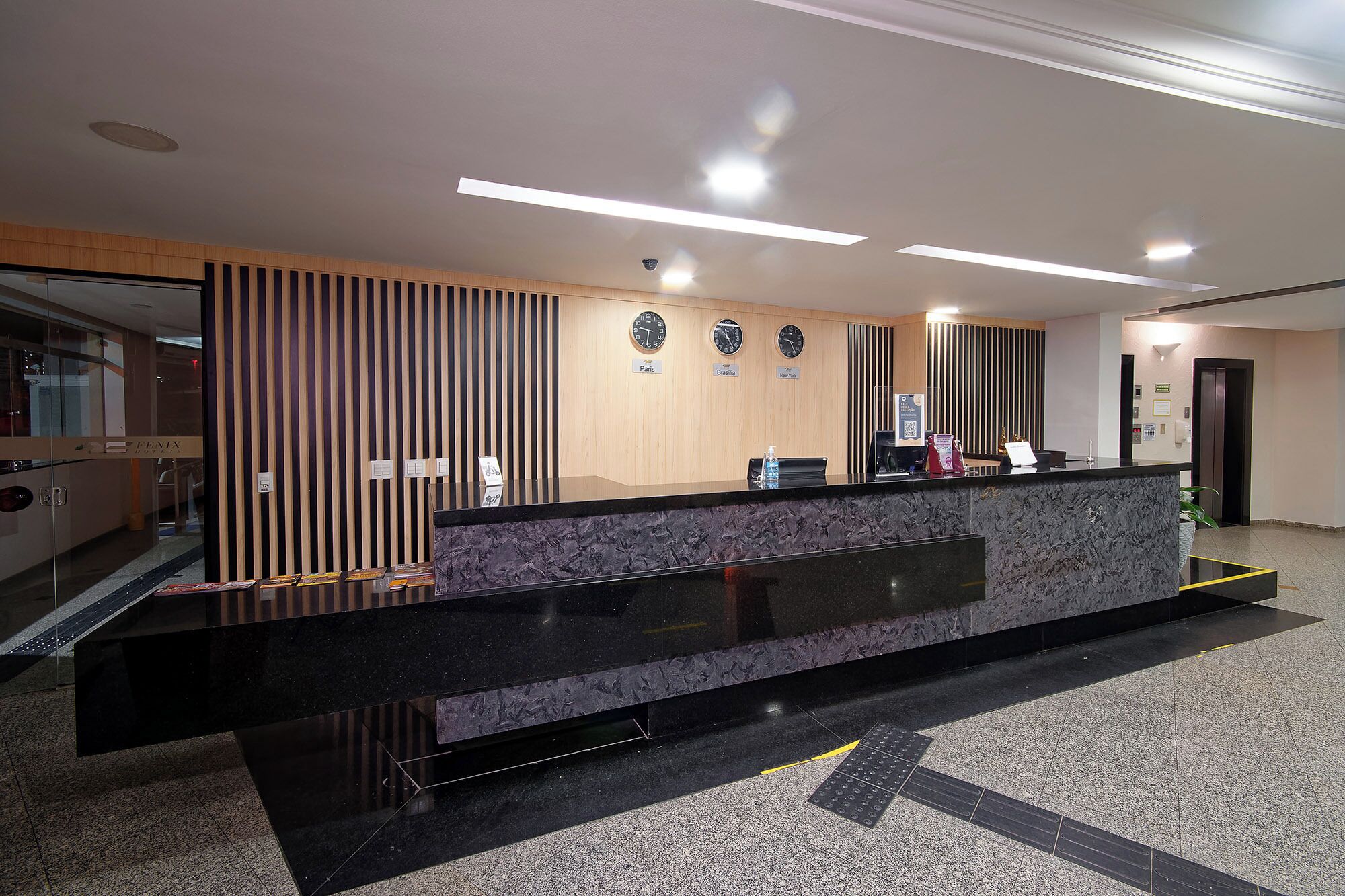Uma área de recepção de hotel com balcão de mármore preto, ripas de madeira verticais e relógios marcando Paris, Brasil e Nova Iorque.