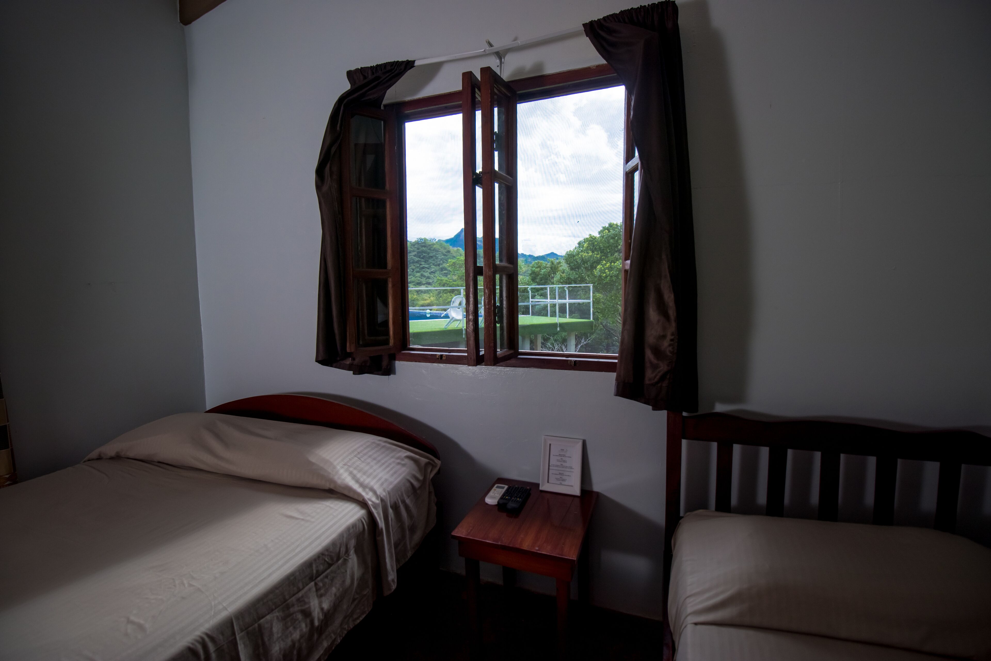 Quarto com duas camas, uma mesinha com um controle remoto e uma janela com cortinas.