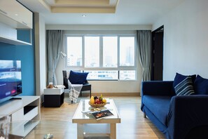 Sala de estar moderna com televisão, mesa de centro com cesta de frutas, sofá e vista da cidade através de grandes janelas.
