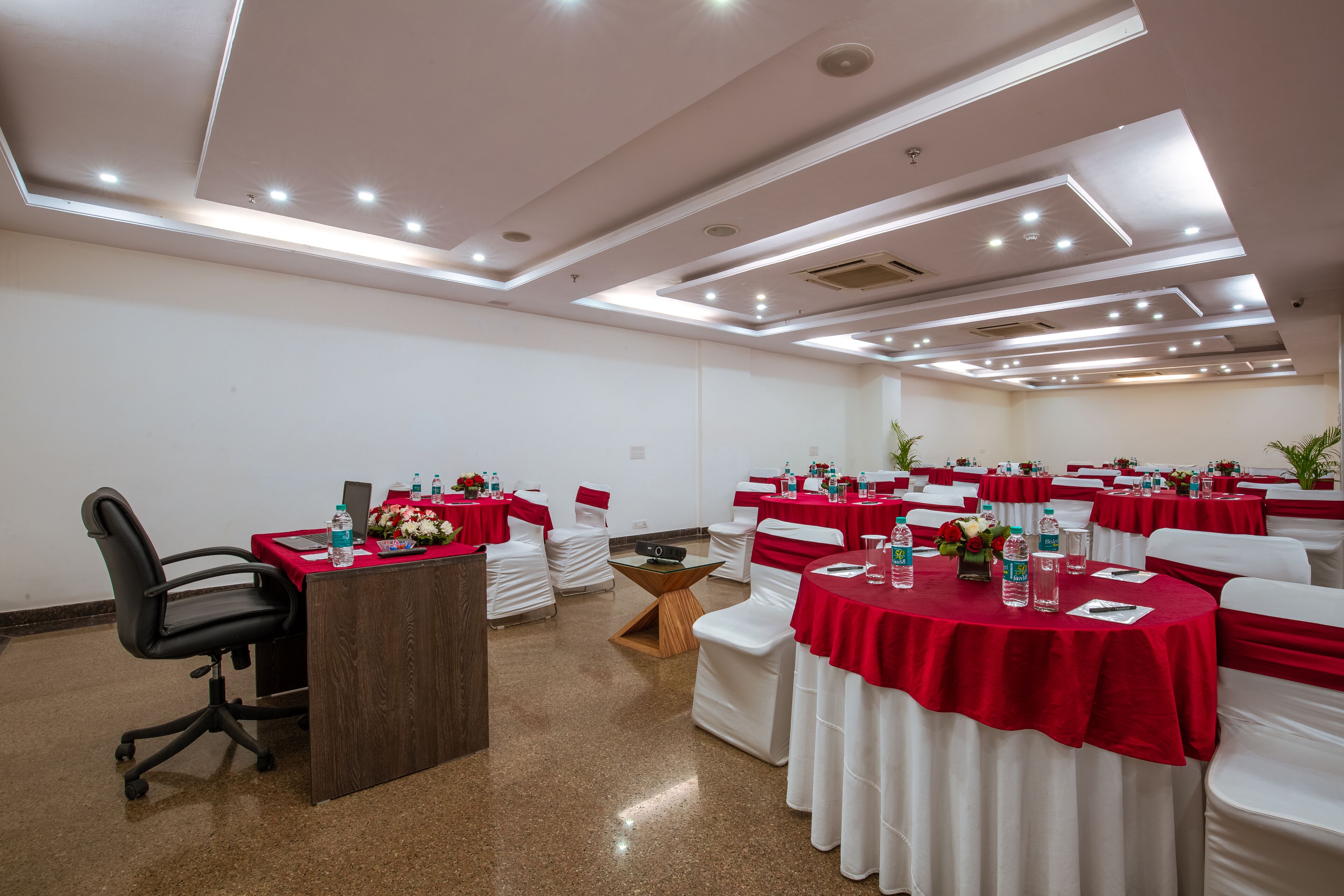 Sala de conferências com mesas redondas, cadeiras brancas e toalhas de mesa vermelhas. Há um púlpito de madeira e uma cadeira de escritório preta.
