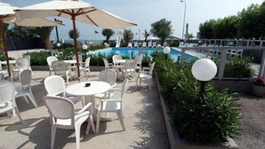 Área de piscina com cadeiras e mesas brancas, guarda-sóis e uma piscina.