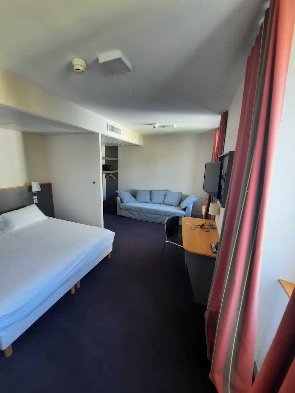 Quarto de hotel com cama, sofá, escrivaninha, televisão e guarda-roupa.
