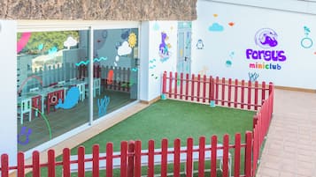 Espaço infantil com cerca vermelha, grama sintética verde e parede decorada com adesivos de animais e o logotipo "Fargus mini club".